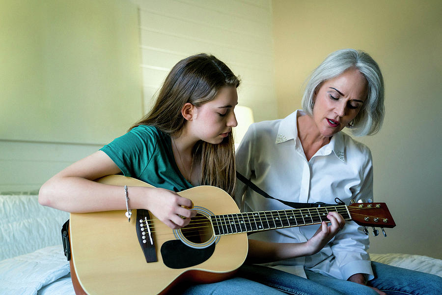 Granddaughter Playing Guitar While Sitting By Grandmother In Bedroom Photograph By Cavan Images
