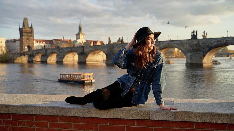Stylish beautiful young woman wearing black hat sitting on Vltava river shore in Prague with ...